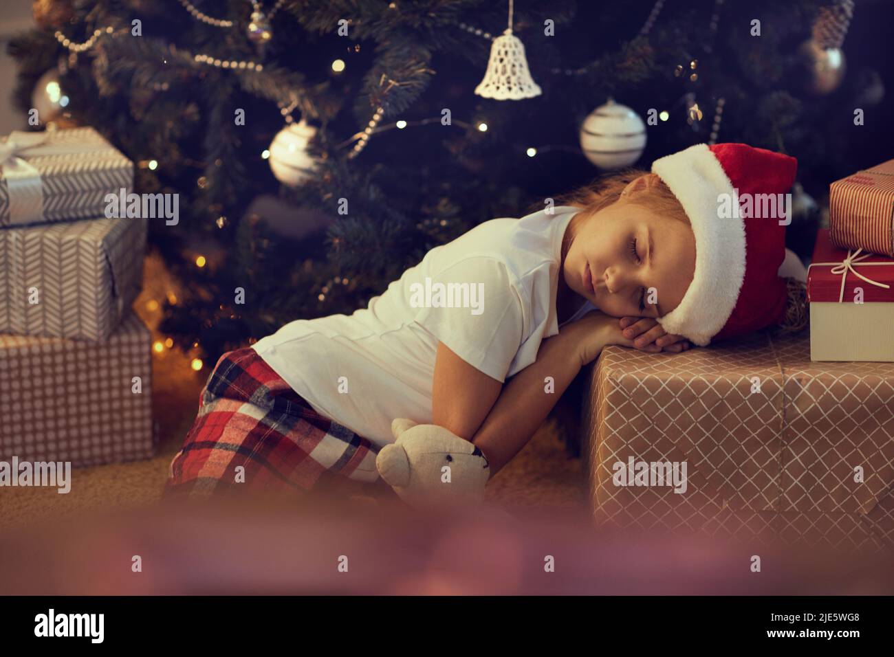 Tired cute girl in pajamas and Santa hat sitting near big gift box and ...