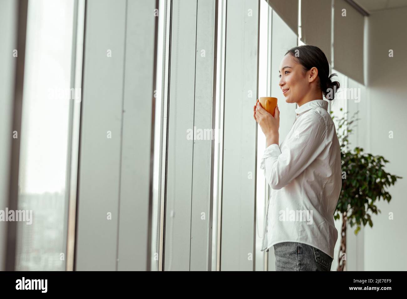 Smiling asian female worker drinking coffee standing near windows at ...