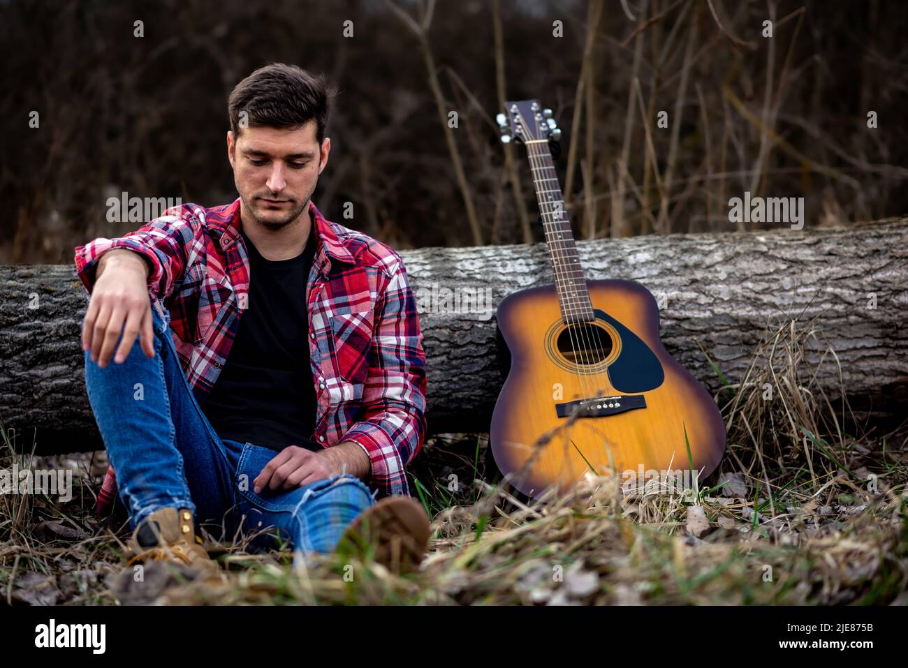 A handsome young man is sitting on ground leaning on a fallen tree next ...