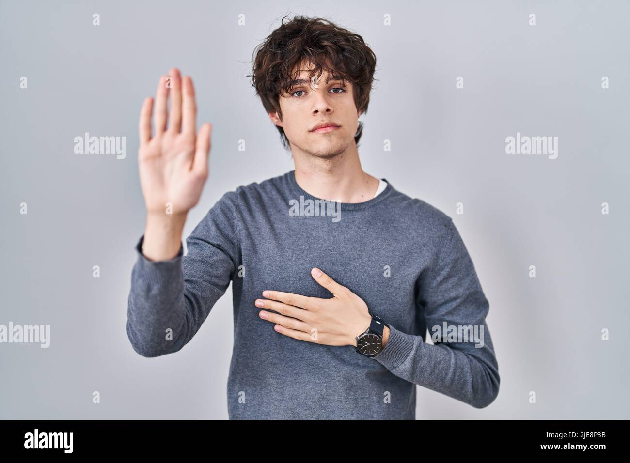 Young man standing over isolated background swearing with hand on chest ...