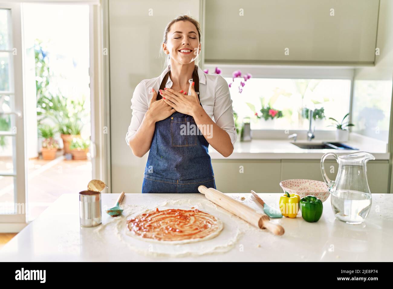 Beautiful blonde woman wearing apron cooking pizza smiling with hands ...