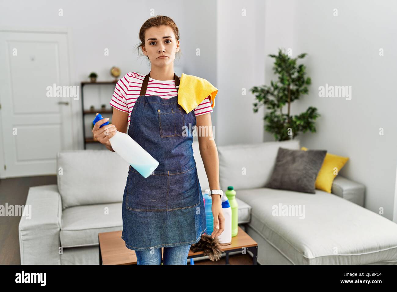 Young brunette woman wearing apron holding cleaning products at home depressed and worry for ...