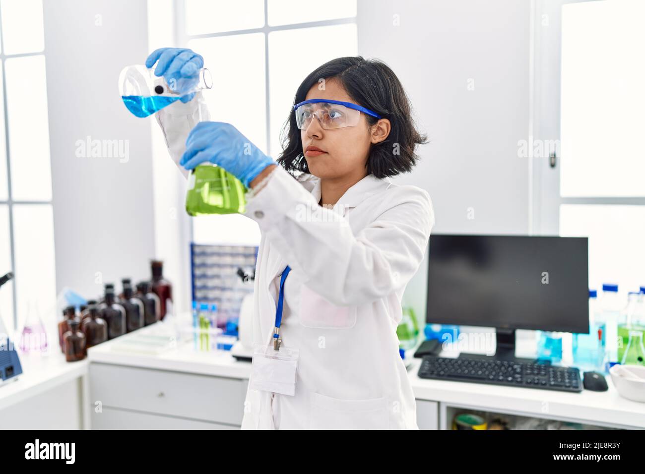 Young latin woman wearing scientist uniform pouring liquid on test tube ...