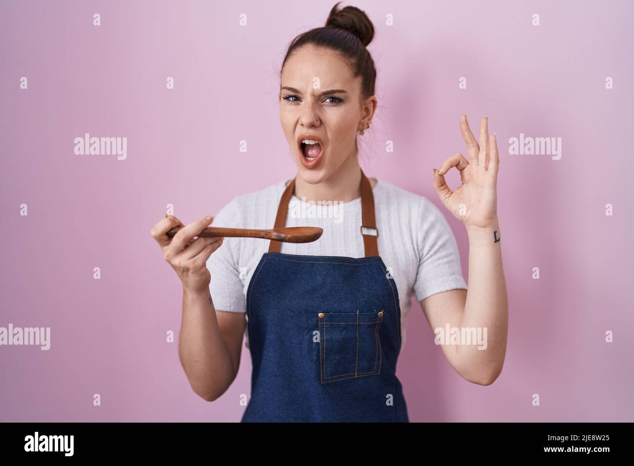 Young hispanic girl wearing professional cook apron holding wood spoon angry and mad screaming ...