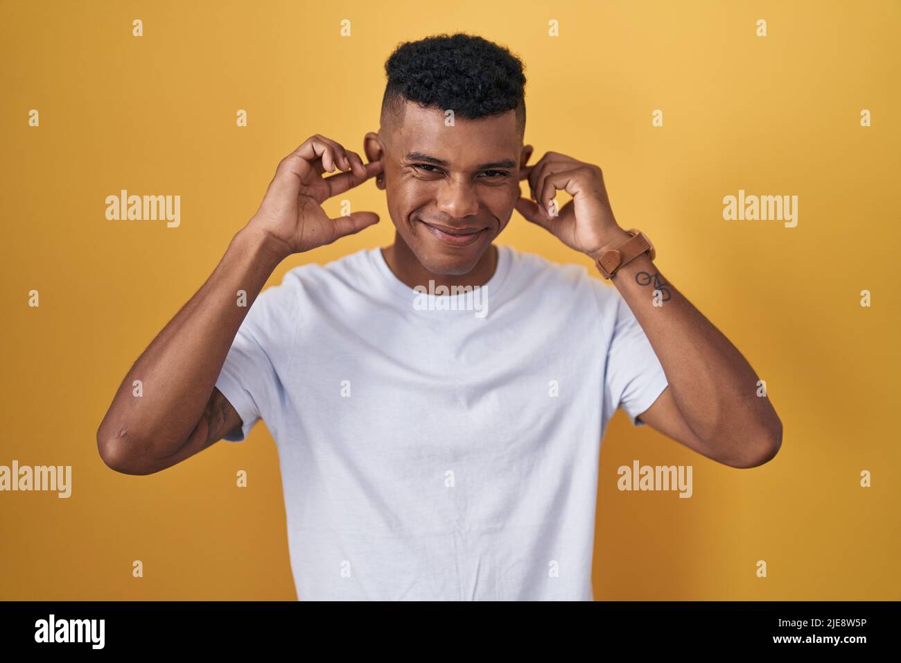 Young hispanic man standing over yellow background covering ears with ...