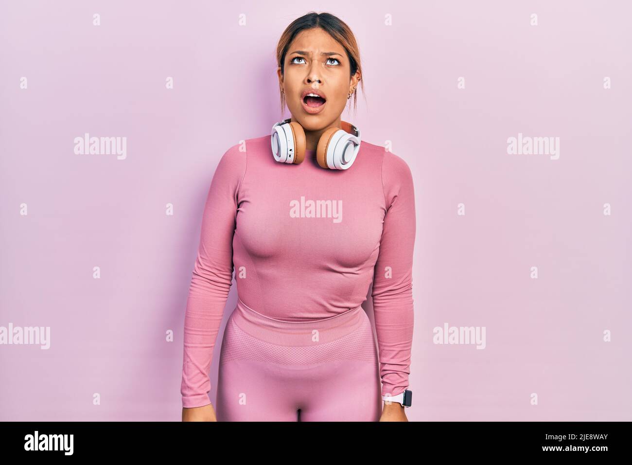 Beautiful hispanic woman wearing gym clothes and using headphones angry ...