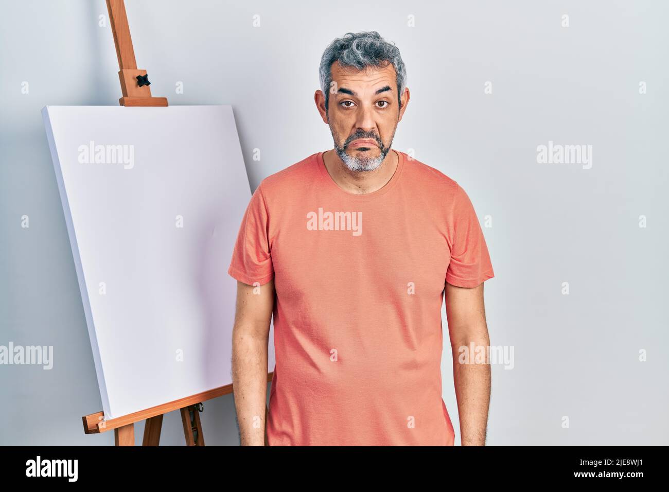 Handsome middle age man with grey hair standing by painter easel stand ...