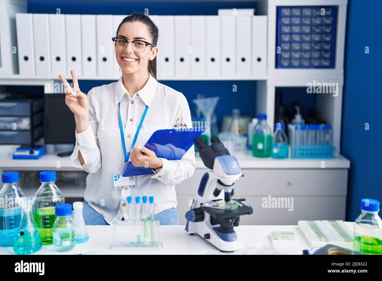 Young brunette woman working at scientist laboratory smiling with happy face winking at the ...