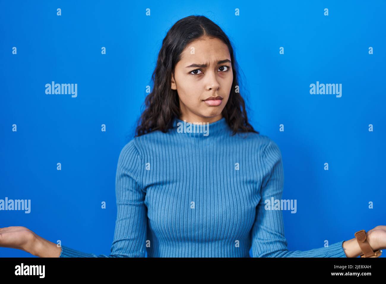 Young brazilian woman standing over blue isolated background clueless ...