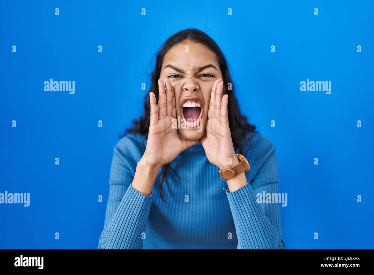 Young brazilian woman standing over blue isolated background shouting ...