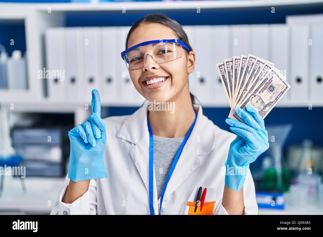 Young brazilian woman working at scientist laboratory holding money surprised with an idea or ...