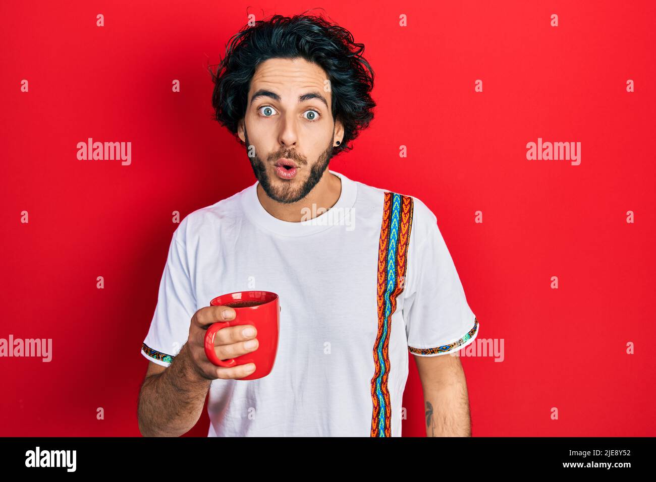 Handsome hispanic man drinking a cup coffee scared and amazed with open ...