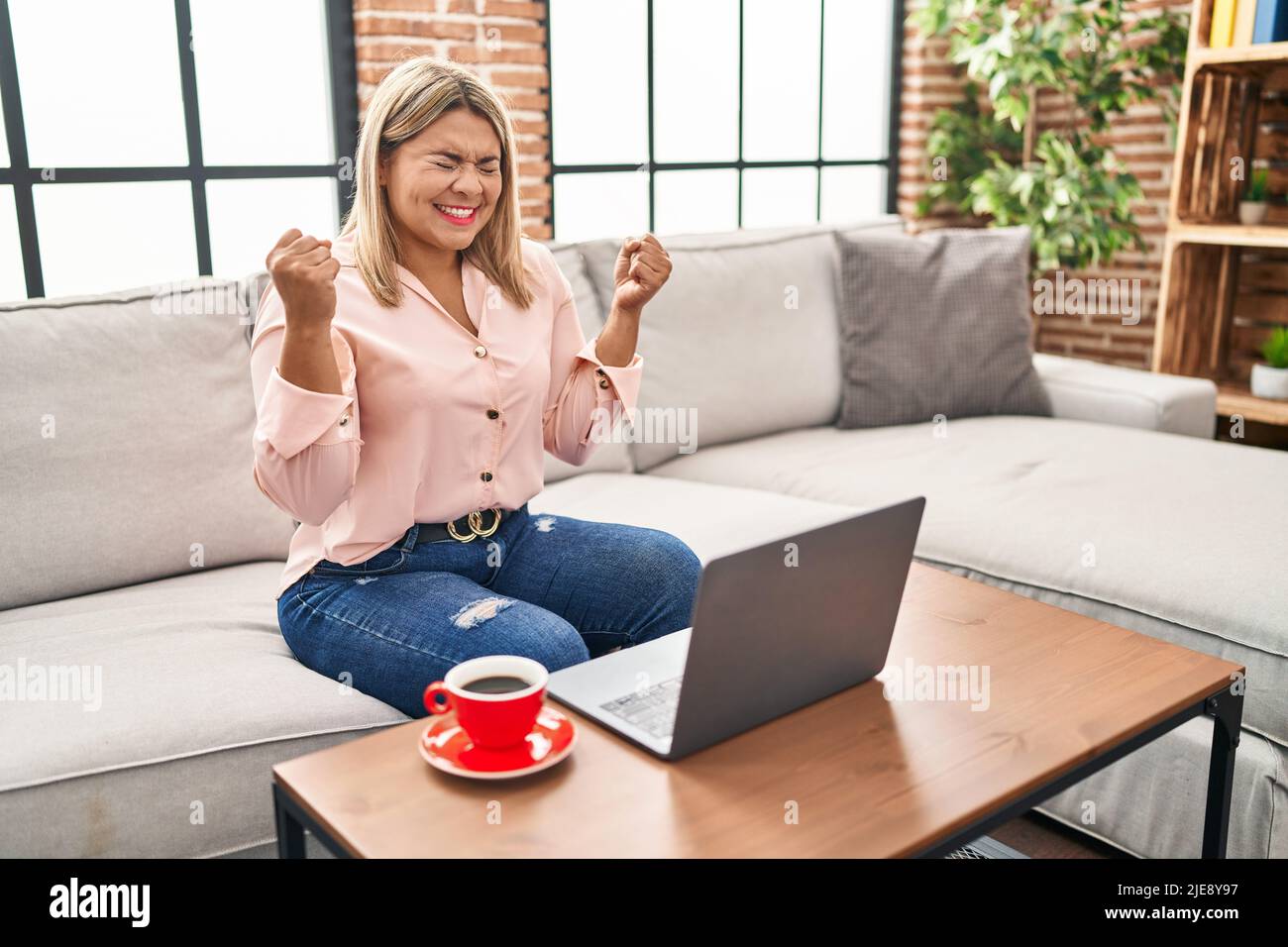 Young hispanic woman using laptop sitting on the sofa at home excited ...