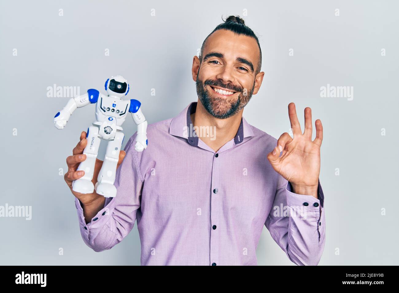 Young hispanic man holding robot toy doing ok sign with fingers ...
