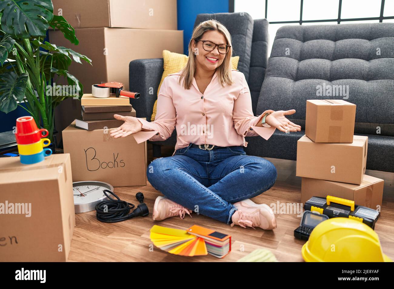 Young hispanic woman moving to a new home sitting on the floor smiling ...