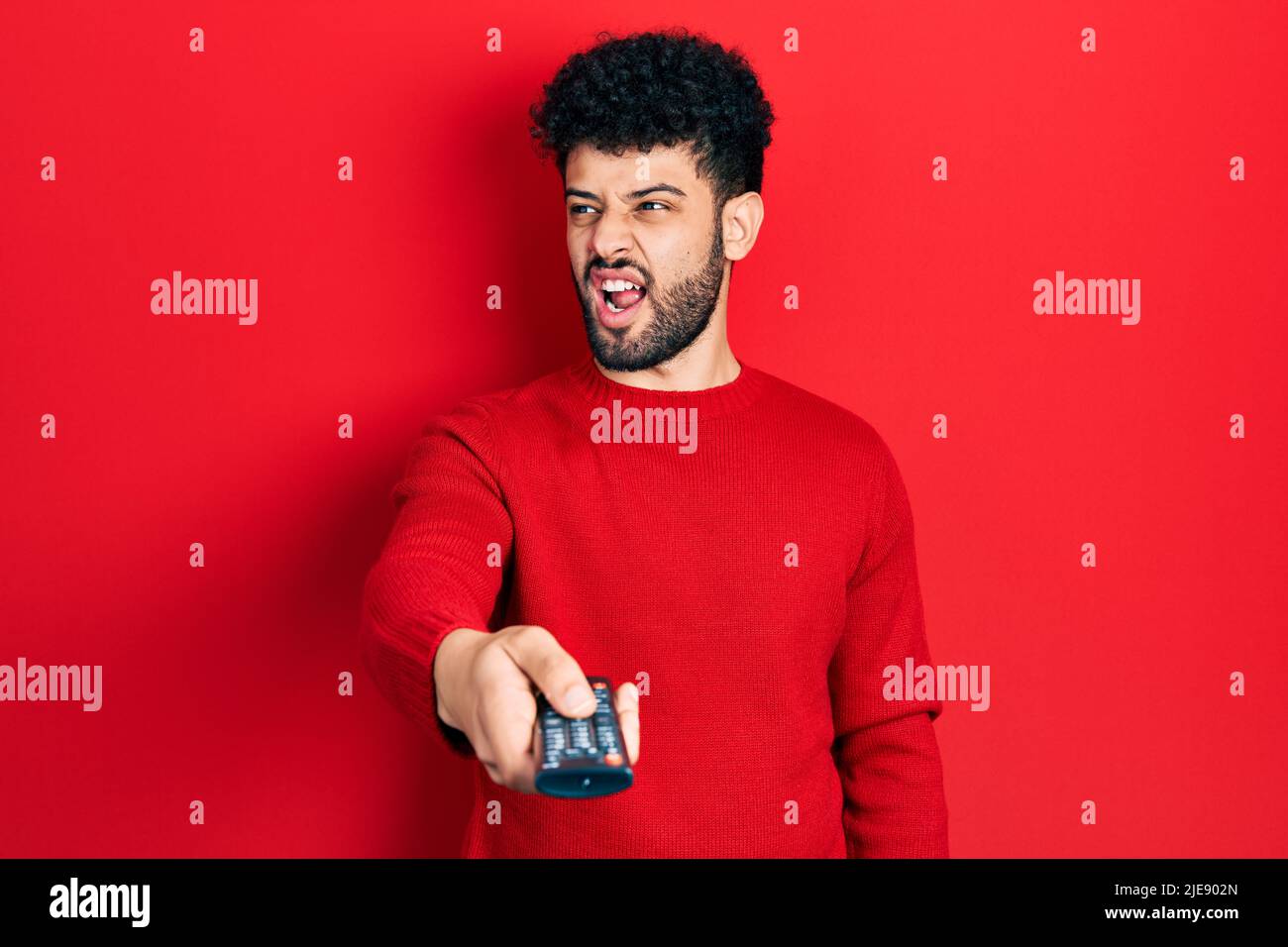 Young arab man with beard holding television remote control angry and ...