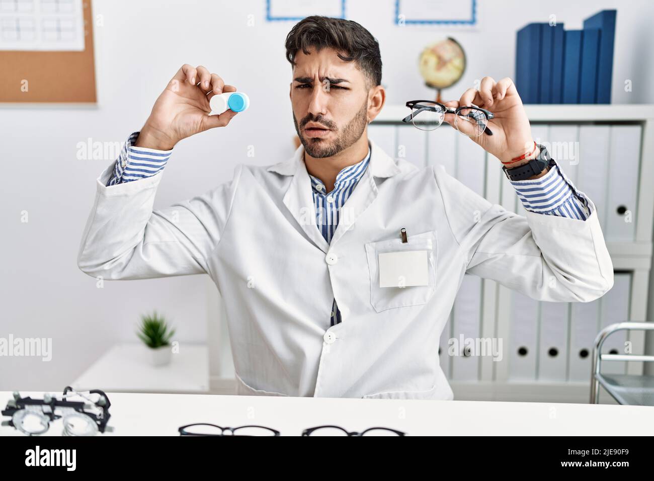 Young optician man holding glasses and contact lenses clueless and ...