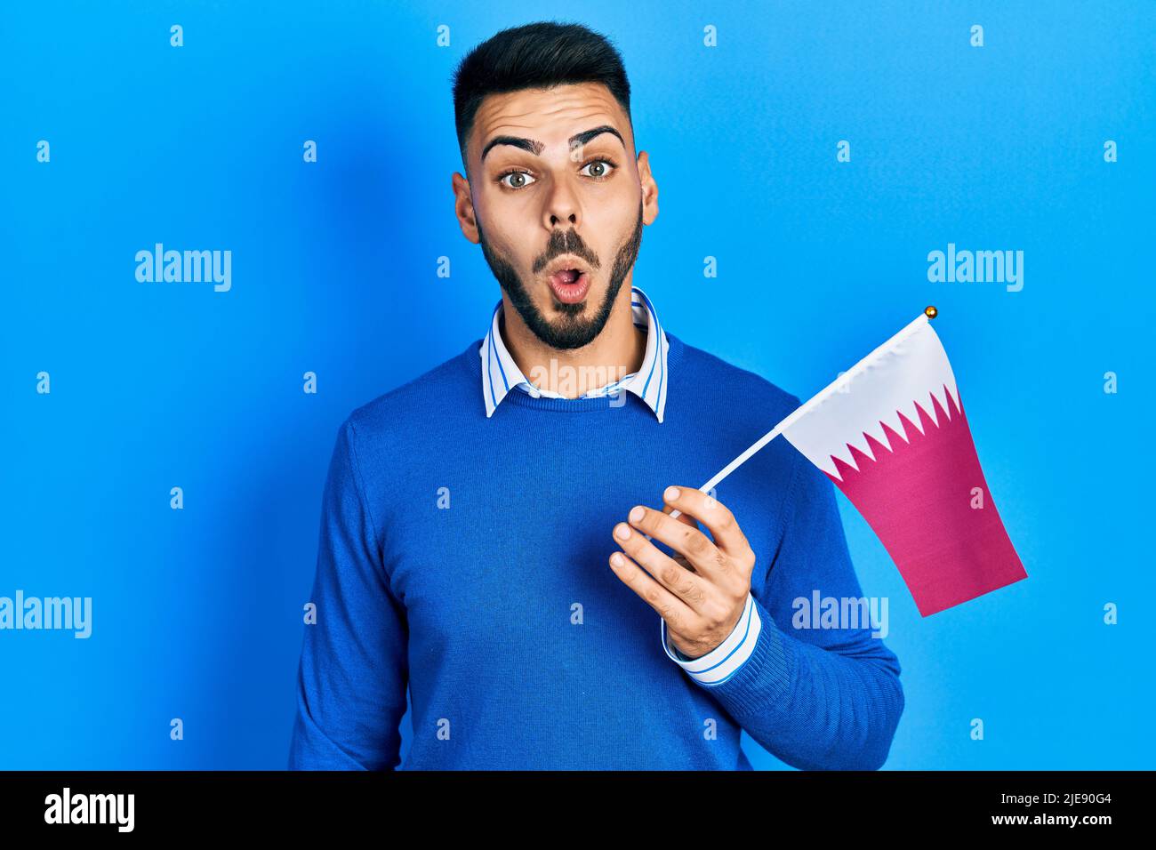 Young hispanic man with beard holding qatar flag scared and amazed with ...