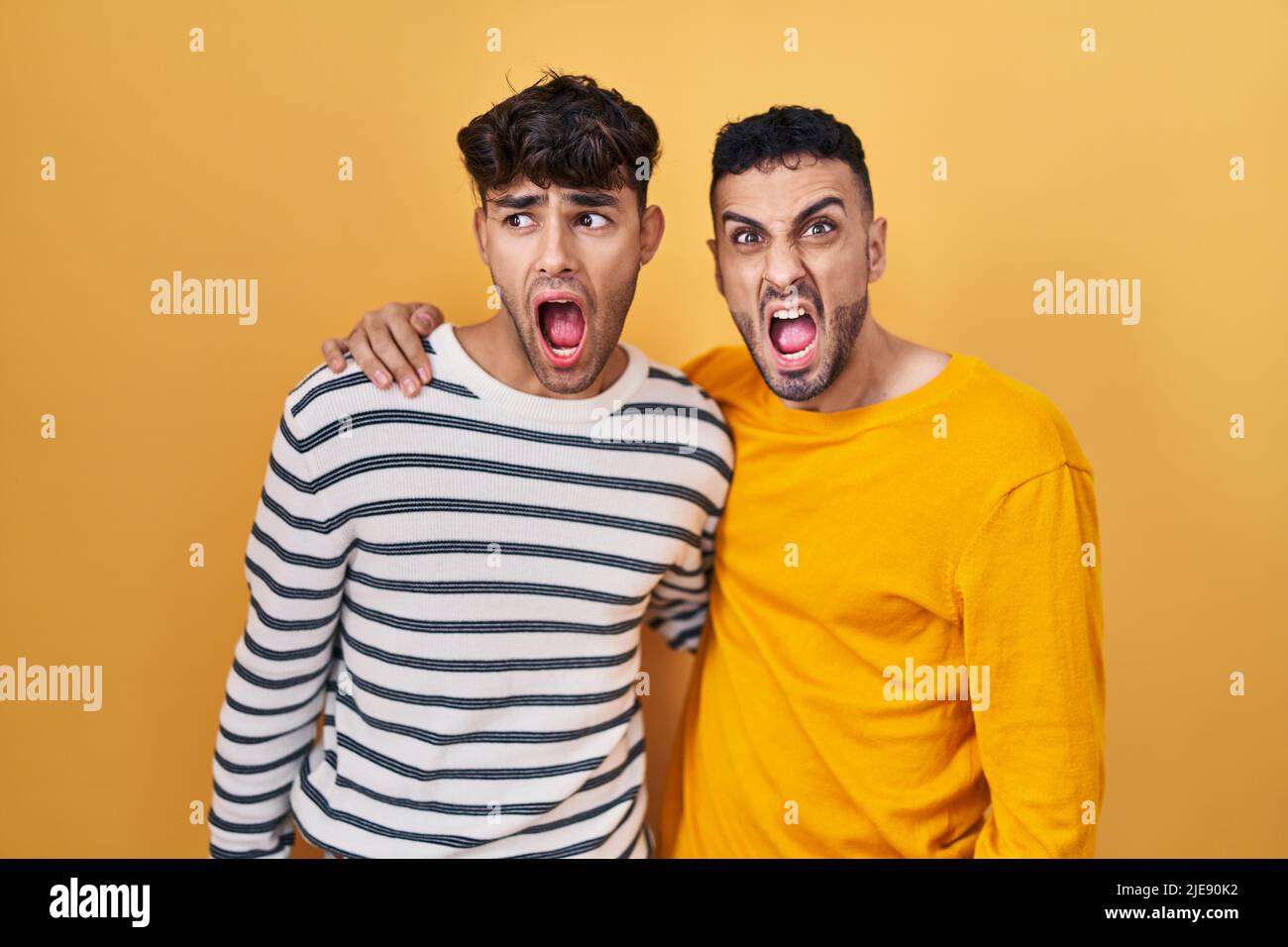 Young hispanic gay couple standing over yellow background angry and mad ...