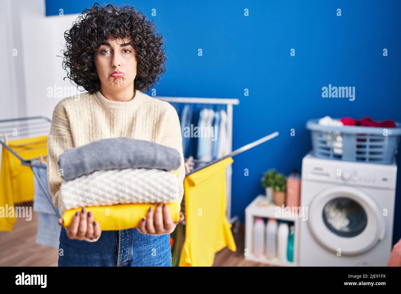 Young brunette woman with curly hair holding clean laundry depressed and worry for distress ...