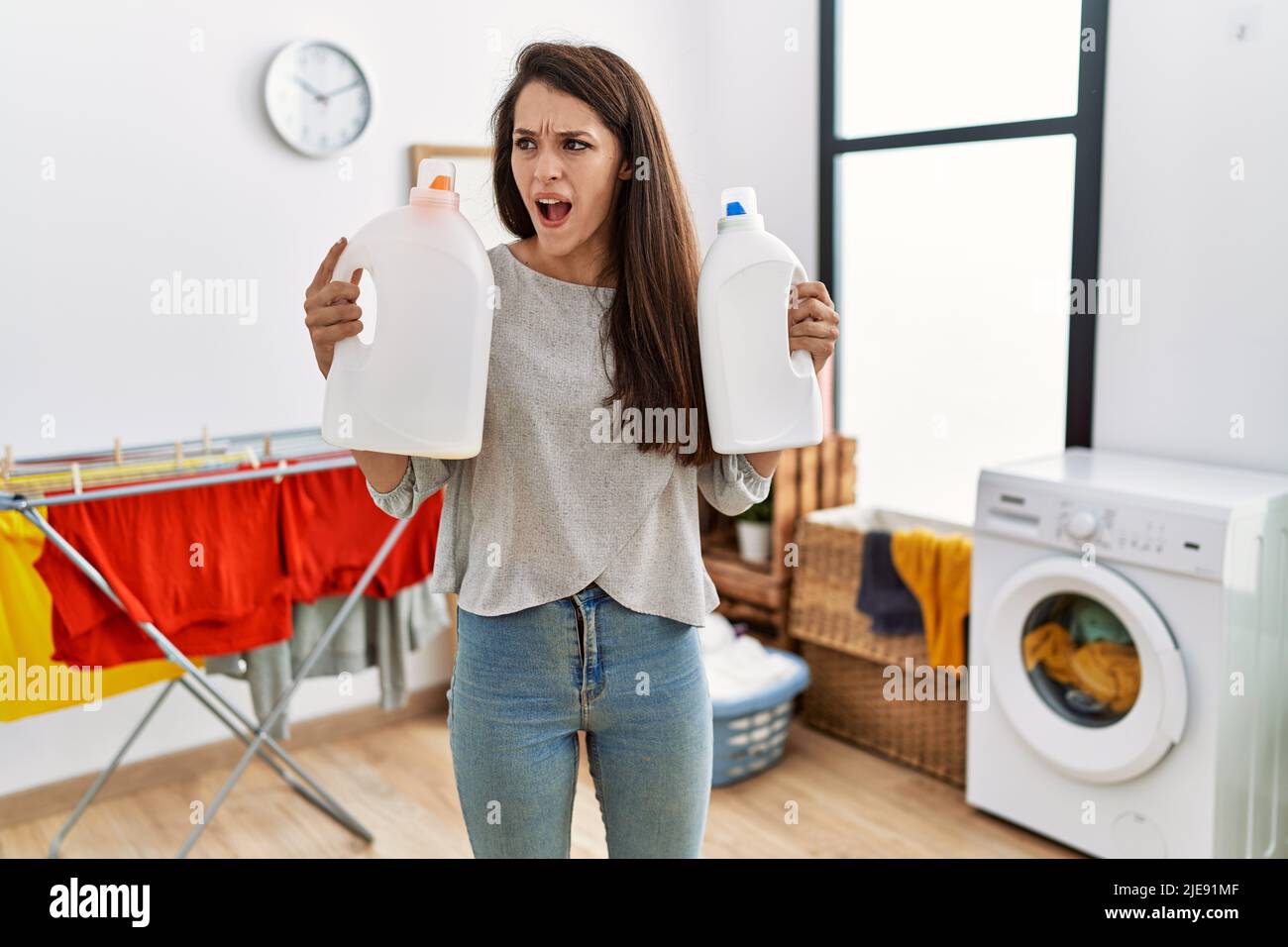 Young brunette woman holding detergent bottle at laundry room angry and mad screaming frustrated ...