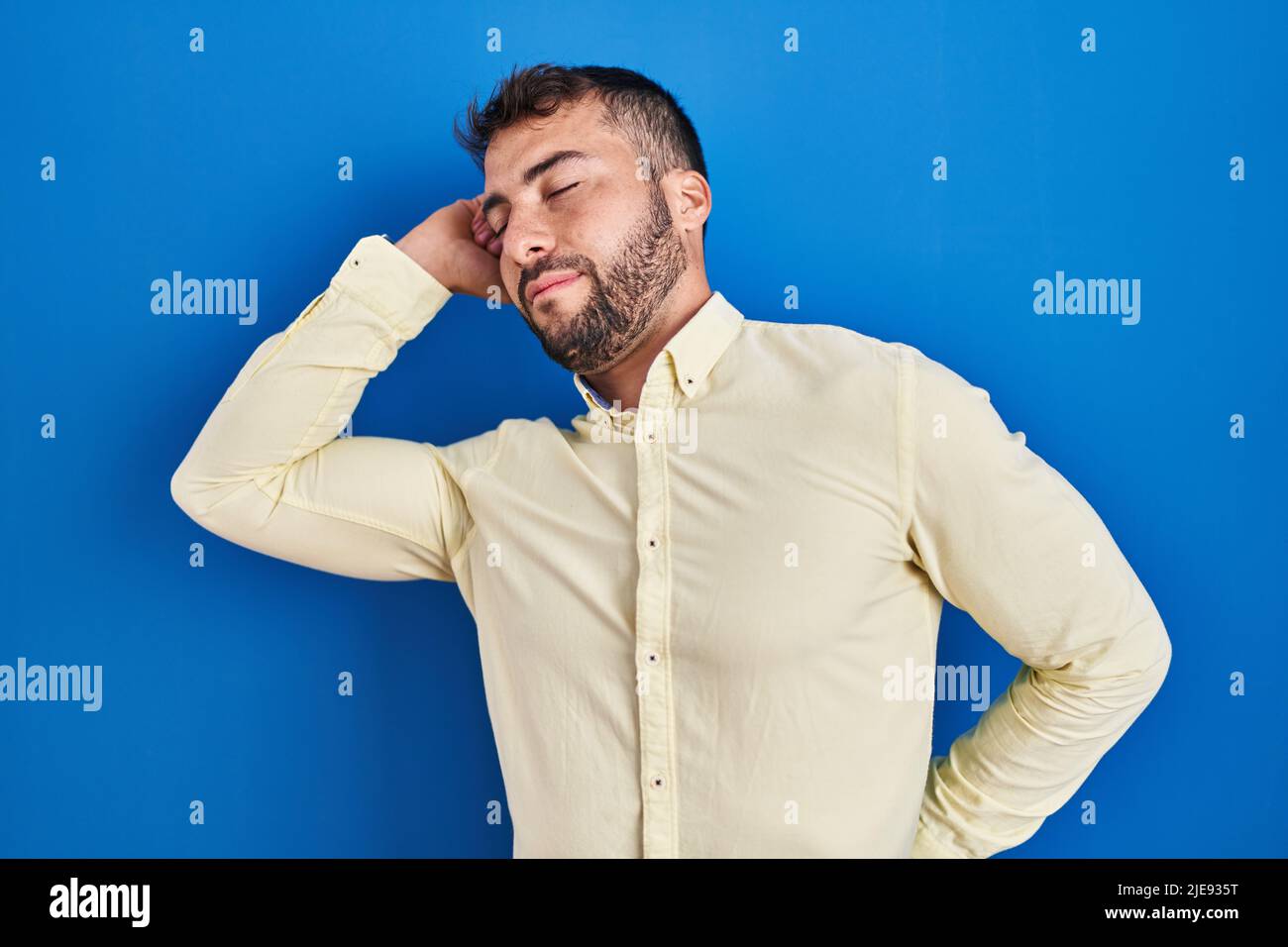 Handsome hispanic man standing over blue background stretching back ...