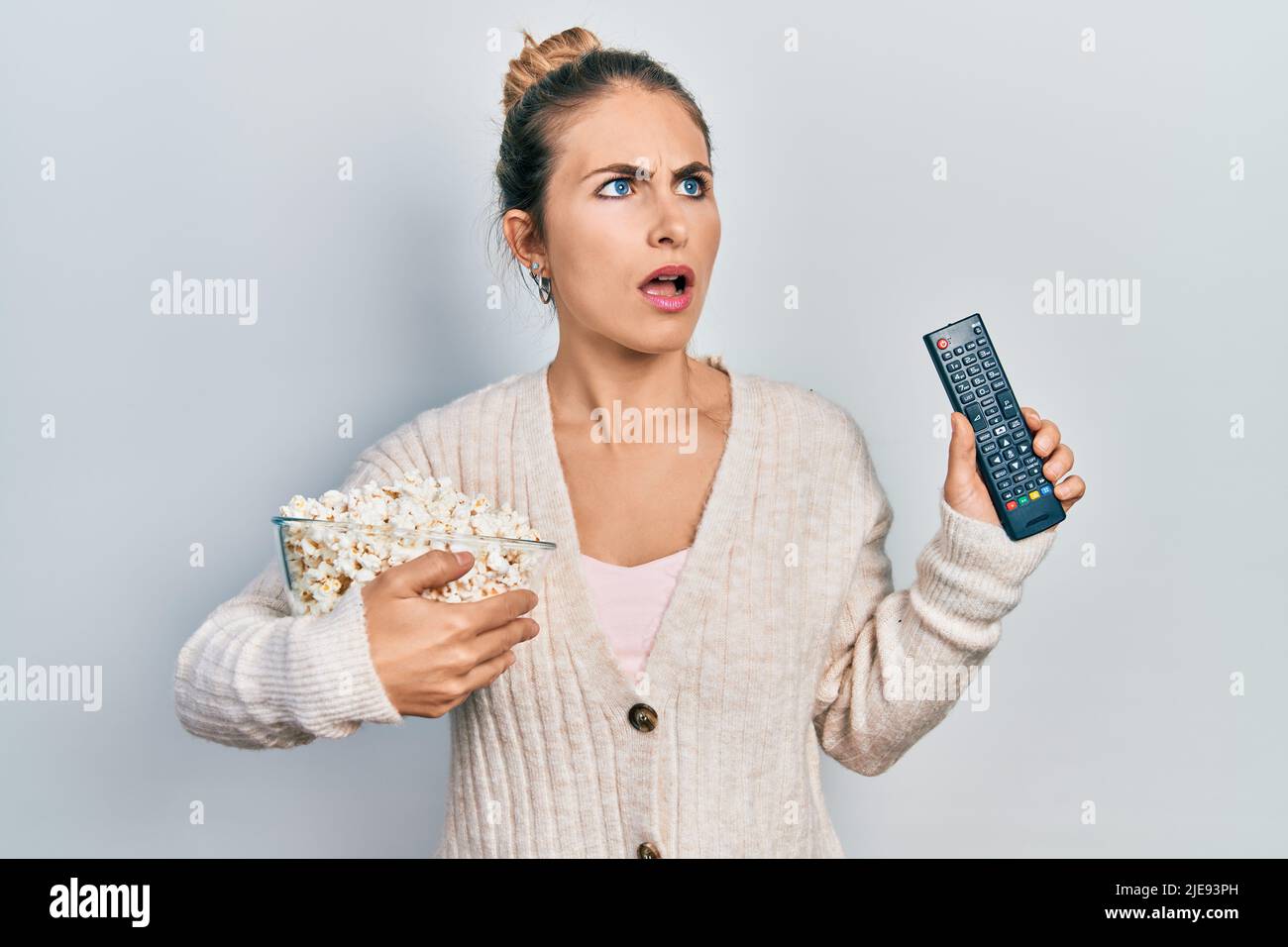 Young caucasian woman eating popcorn using tv control angry and mad ...