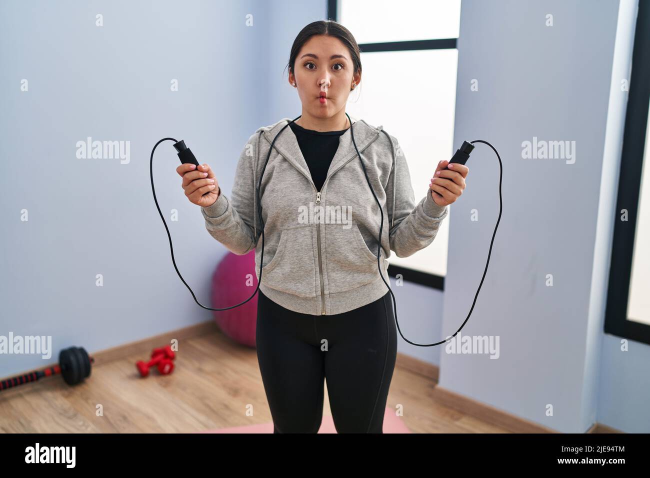 Young latin woman jumping with skipping rope making fish face with ...