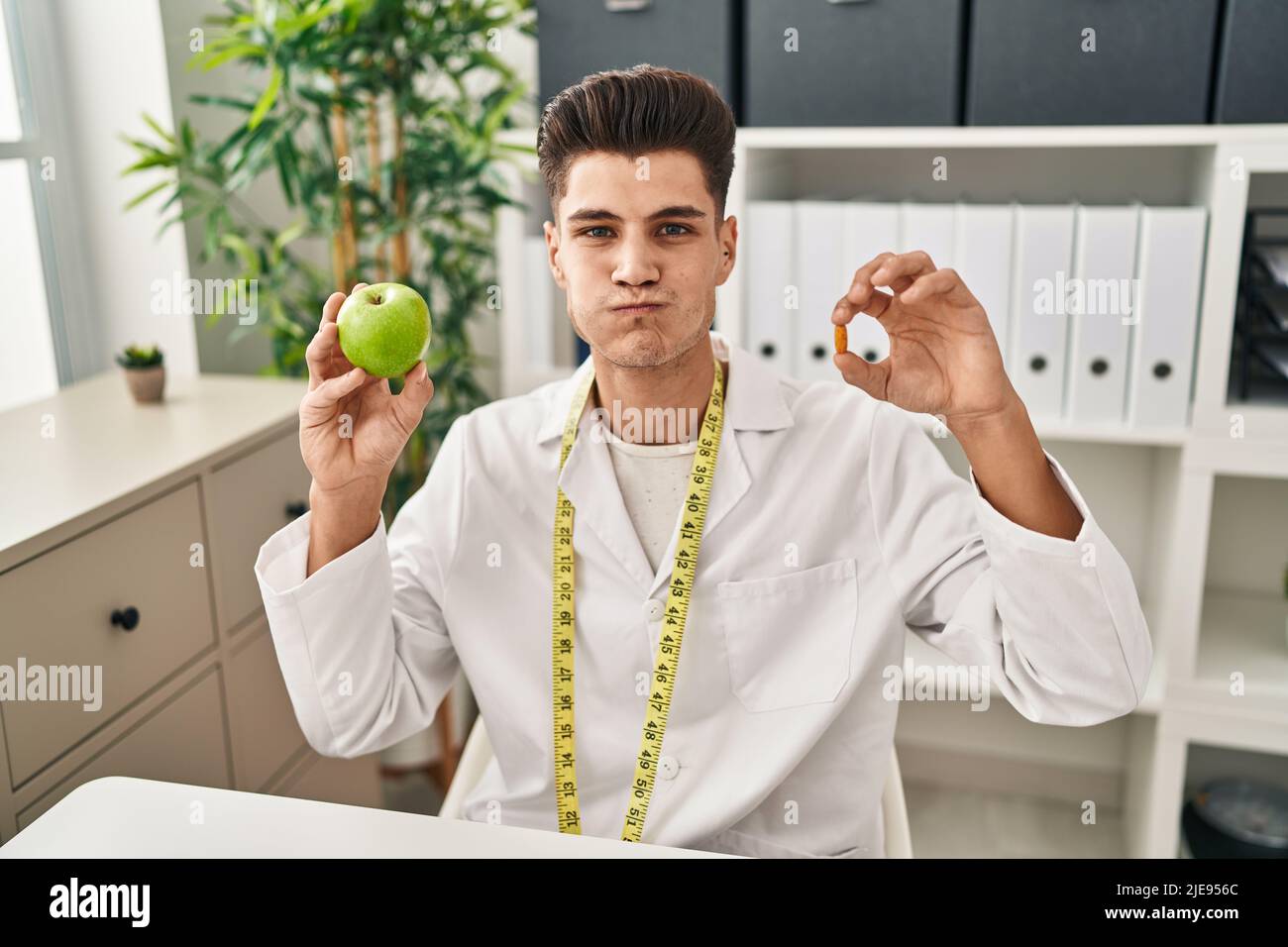 Young hispanic doctor man holding pills for fat loss puffing cheeks ...