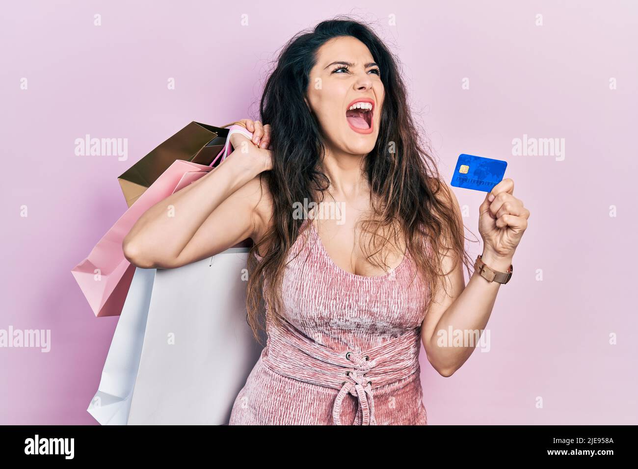 Young hispanic woman wearing santa claus costume holding shopping bags ...