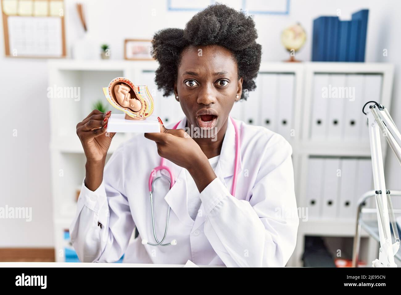 African doctor woman holding anatomical model of female uterus with fetus scared and amazed with ...
