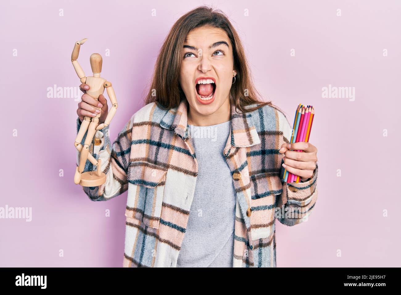 Young caucasian girl holding small wooden manikin and pencils angry and ...