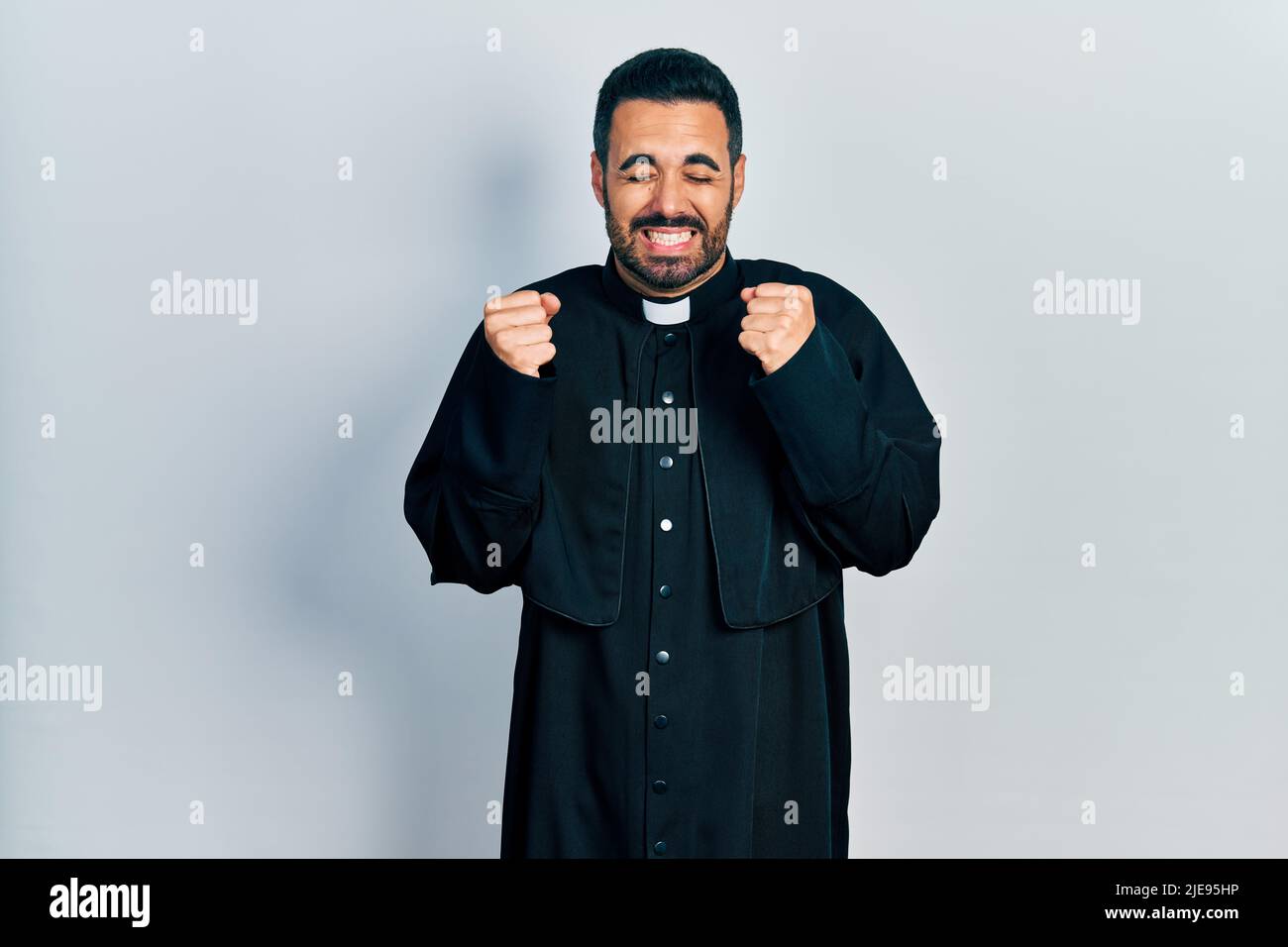 Handsome hispanic man with beard wearing catholic priest robe excited ...