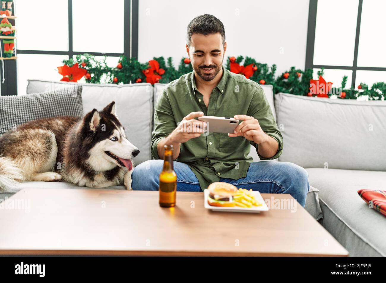 Young hispanic man make photo to hamburger sitting on sofa with dog by ...