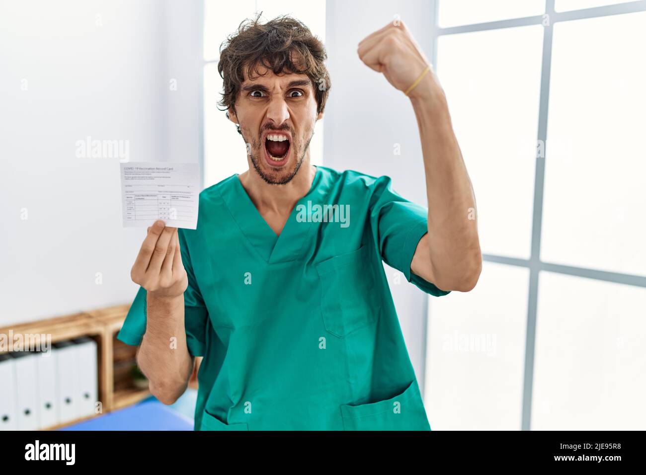 Young hispanic doctor man holding covid record card angry and mad ...