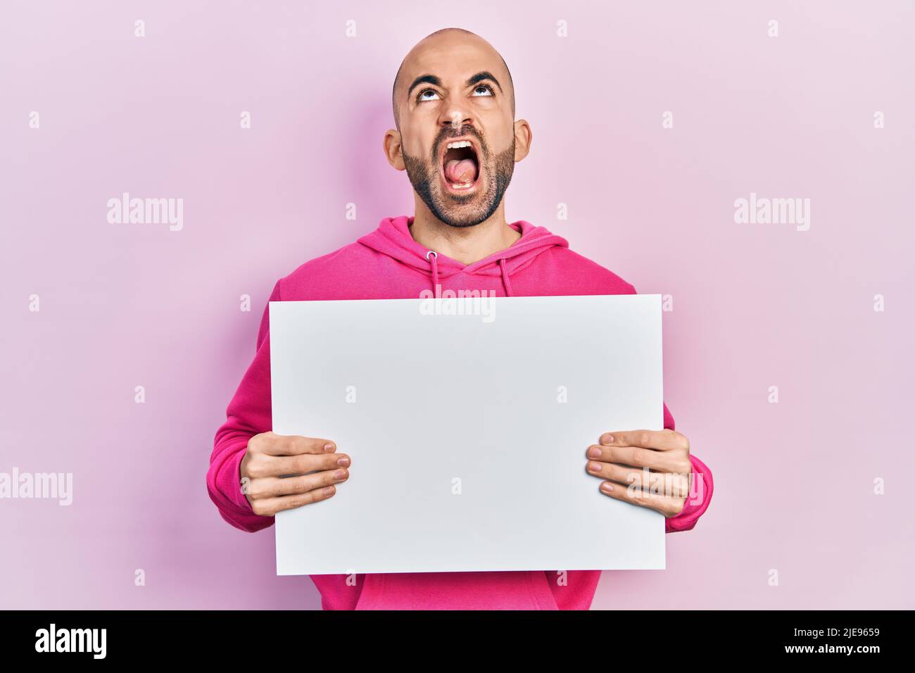 Young bald man holding blank empty banner angry and mad screaming ...