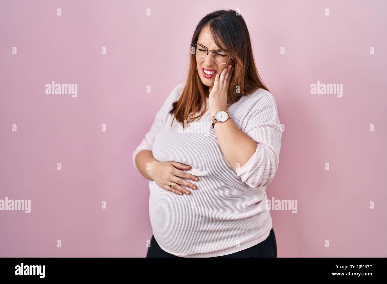 Pregnant woman standing over pink background touching mouth with hand ...