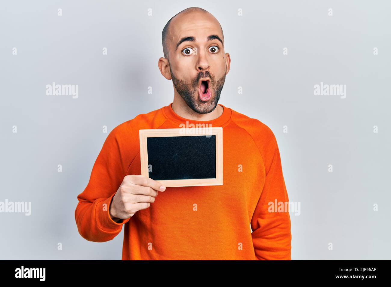 Young bald man holding blackboard scared and amazed with open mouth for ...