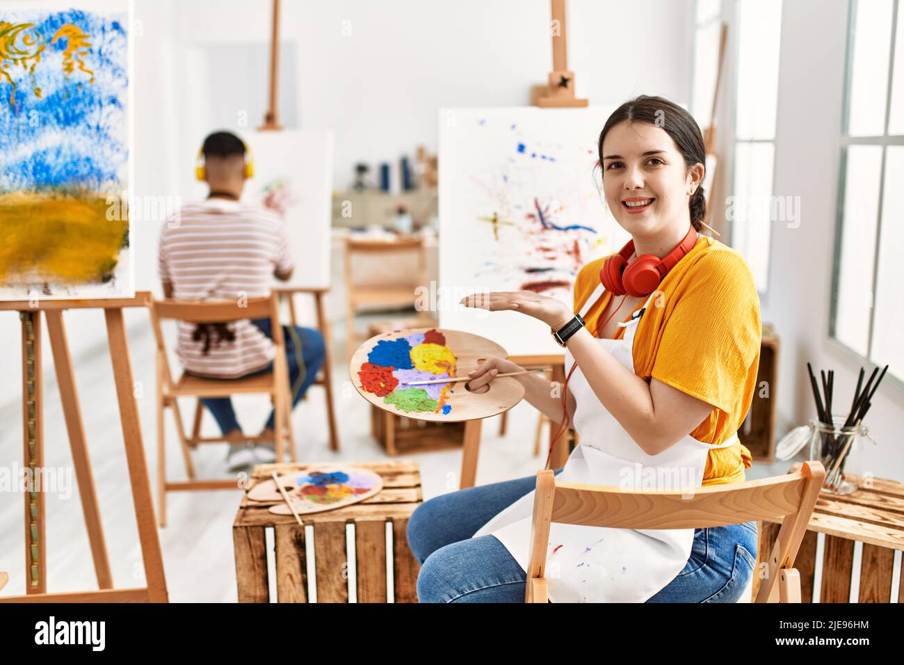 Young artist woman painting on canvas at art studio pointing aside with ...