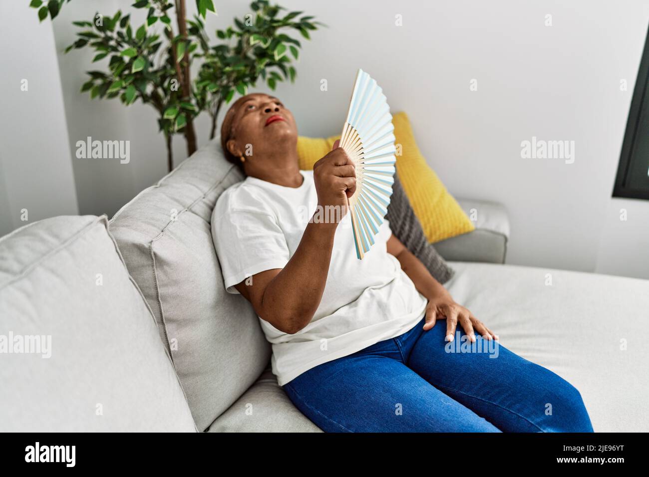 Senior african american woman using hand fan sitting on sofa at home ...