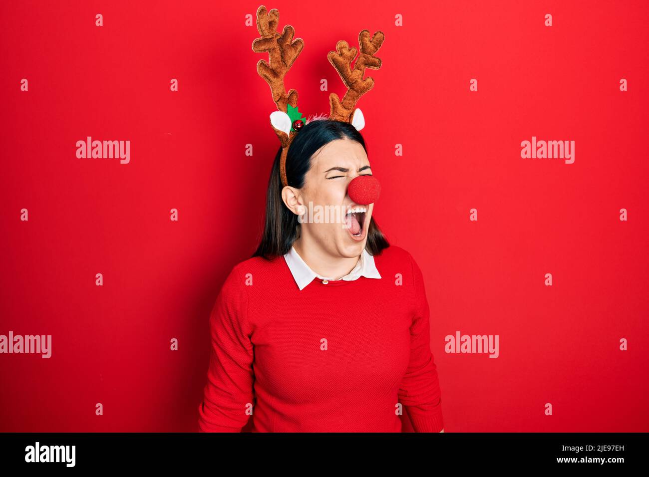 Young hispanic woman wearing deer christmas hat and red nose angry and ...