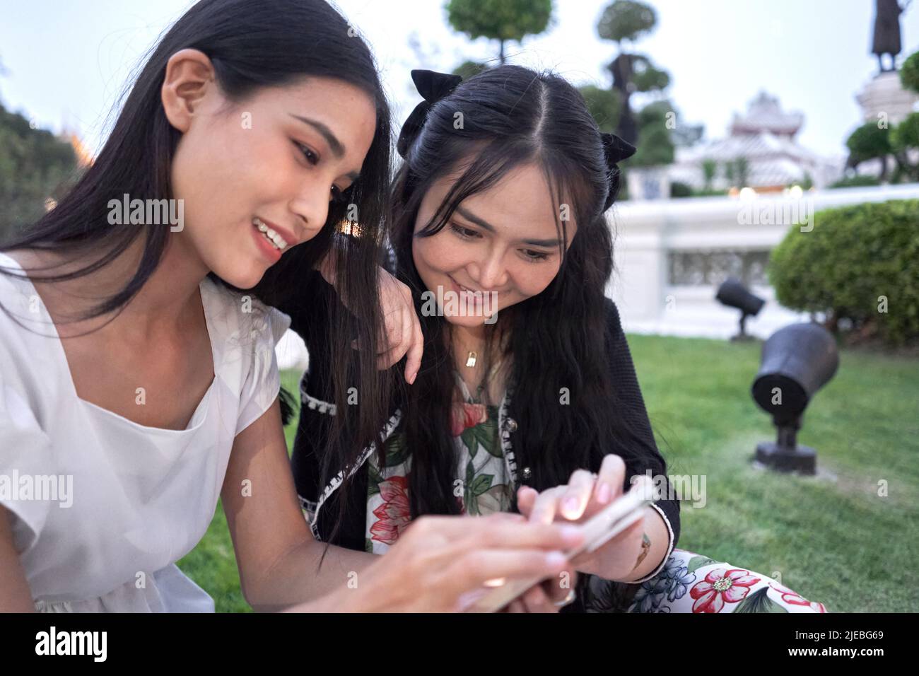 Two smiling friends using the mobile together in a garden of a buddhist ...