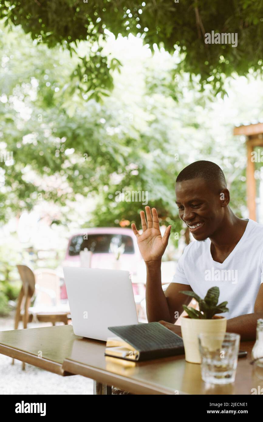 Smiling and glad multiracial man with laptop, raising arm, greet ...