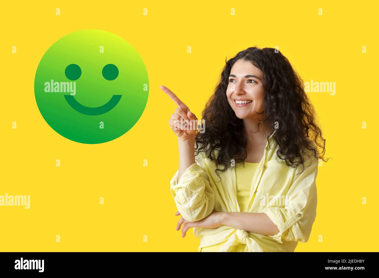 Beautiful young woman pointing at smiling emoticon on yellow background ...
