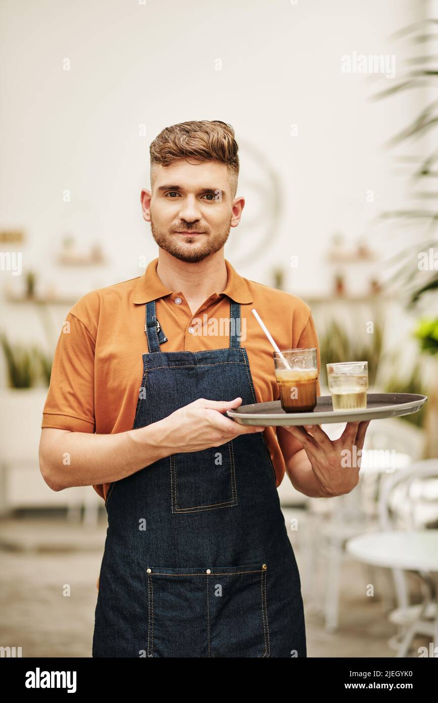 Portrait of positive waiter holding tray of fresh water and iced coffee ...