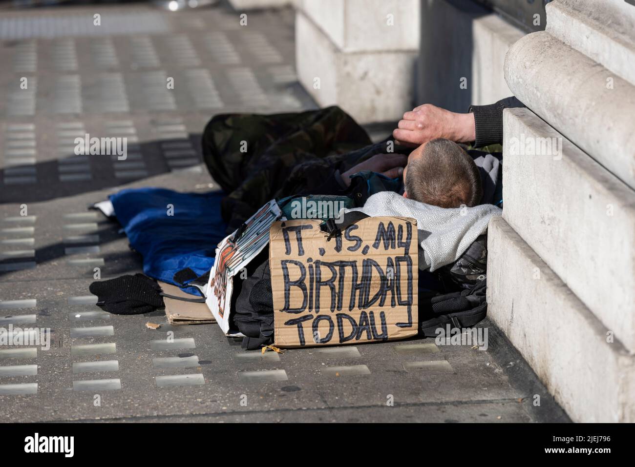 Homeless man lying on the street on a makeshift bed with a sign stating ...