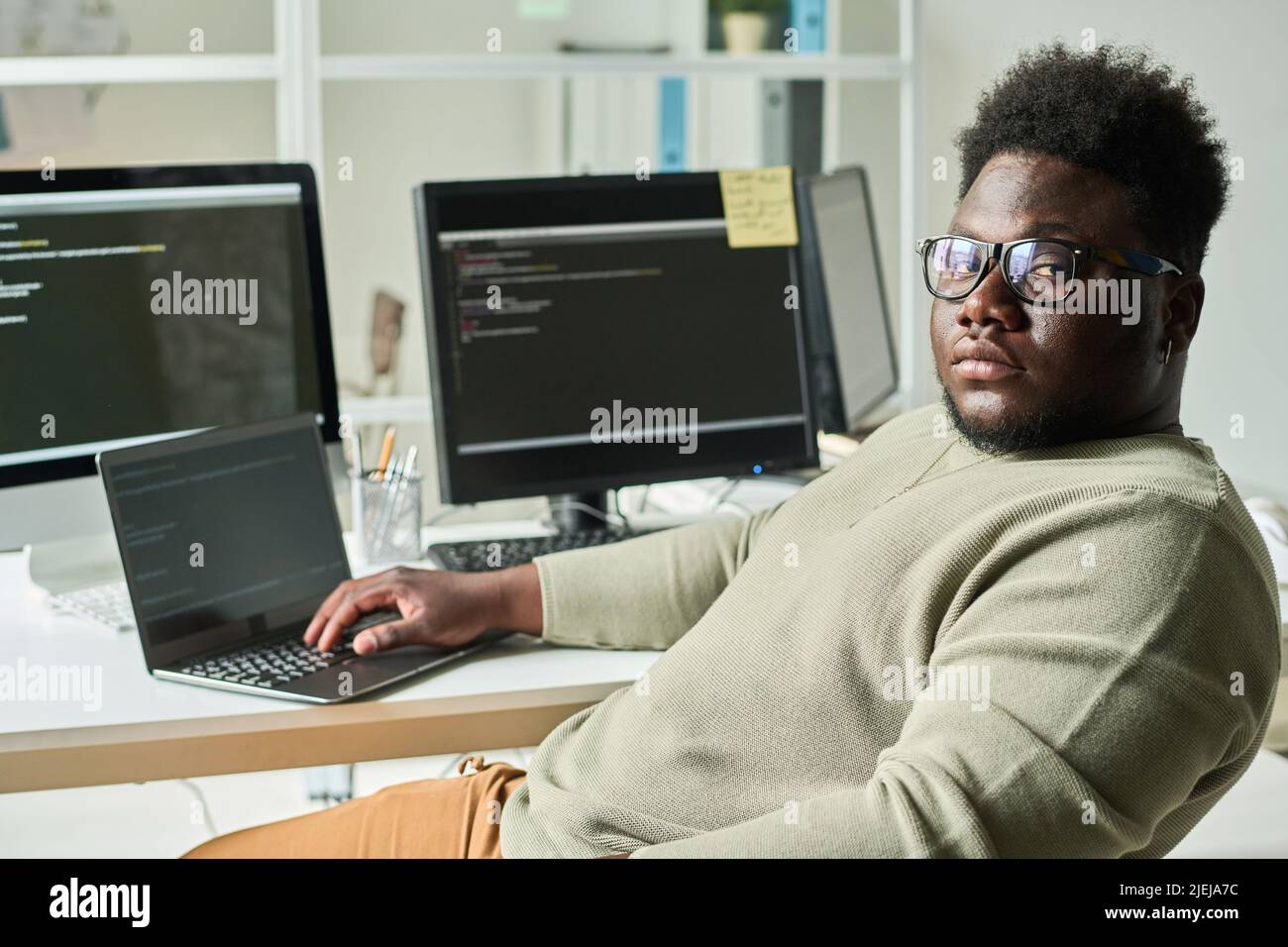 Serious African Programmer Looking At Camera While Sitting Near Computer Monitor With Script On