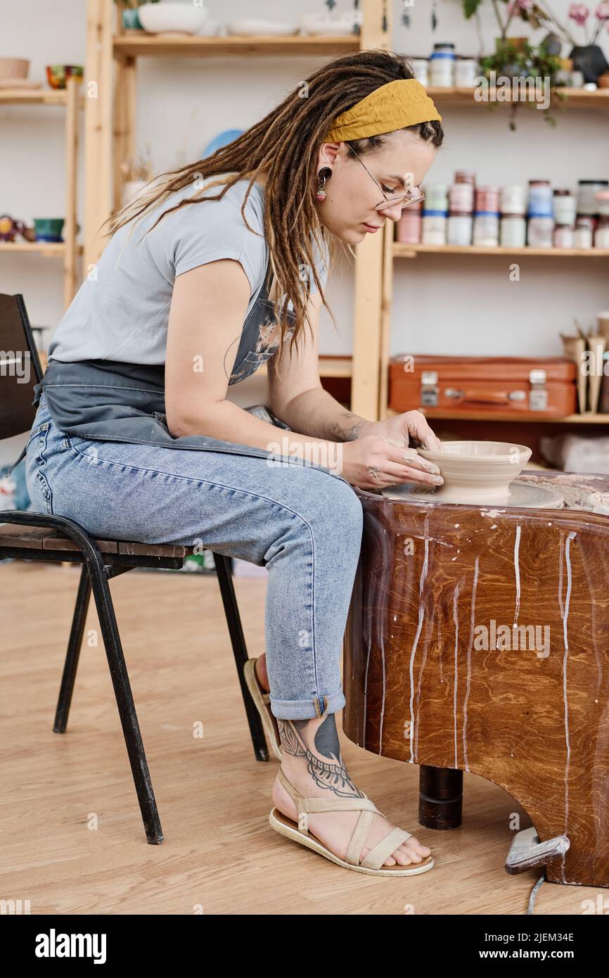 Young creative female artisan sitting by rotating pottery wheel and ...