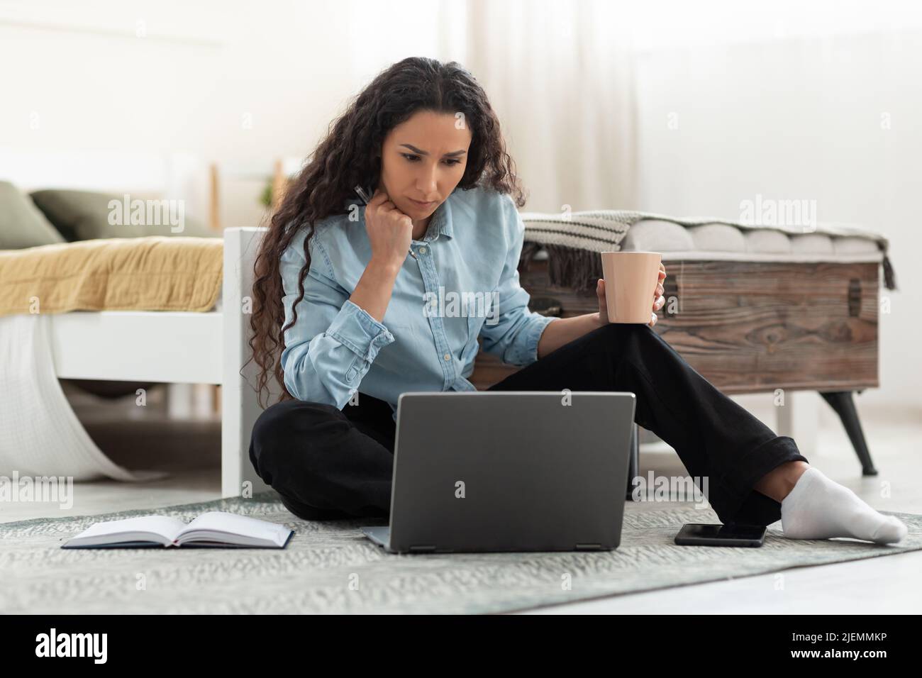 Thoughtful millennial female using laptop pc, sitting on floor with ...
