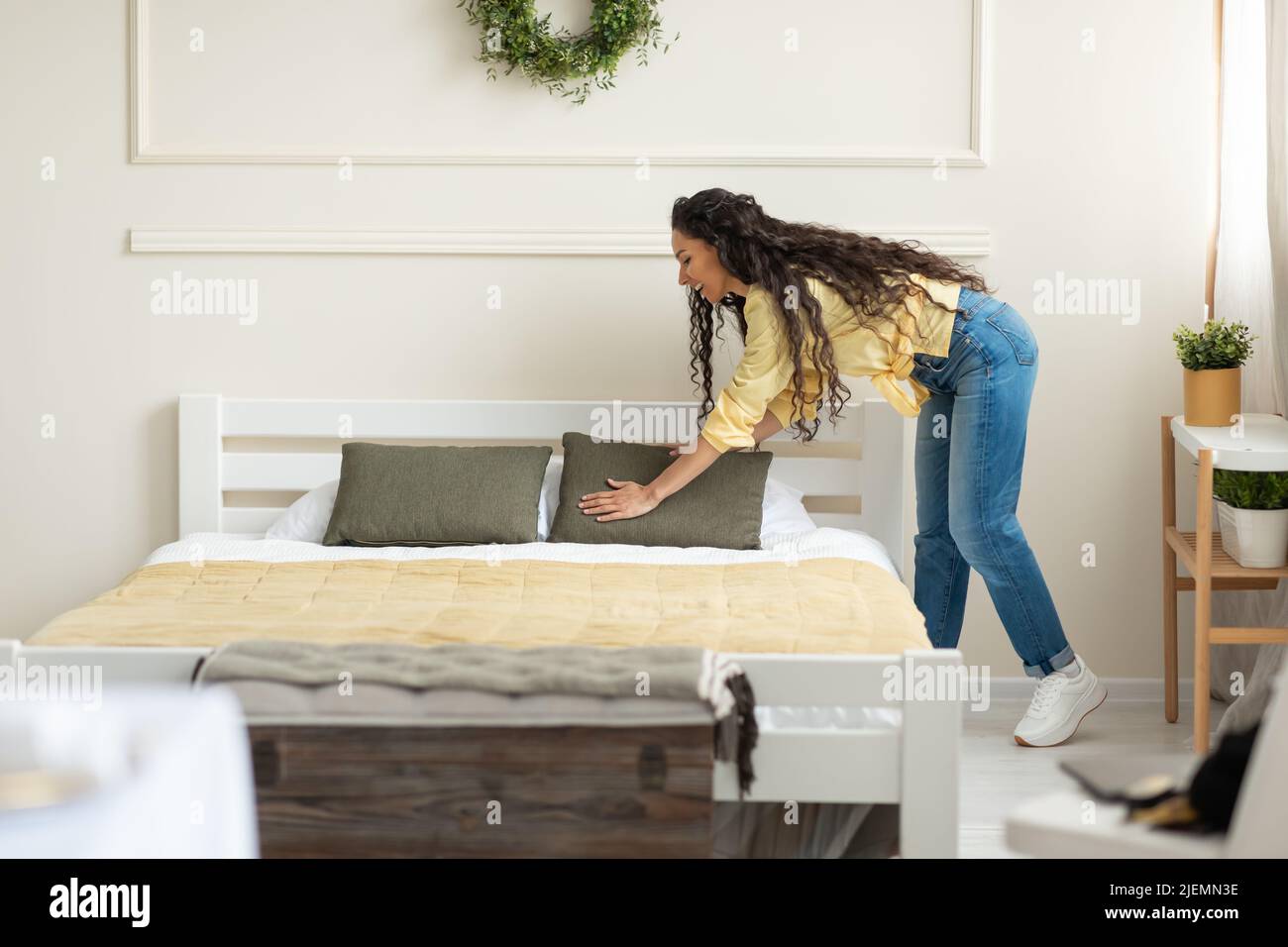 Cheerful young lady making bed at home, arranging pillows, doing ...